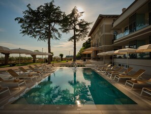 Uma área de piscina com espreguiçadeiras e guarda-sóis, cercada por árvores e um edifício com varandas.