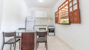 Uma cozinha compacta com armários brancos, forno embutido e geladeira. Há uma mesa de jantar de vidro com cadeiras e uma cadeira de vime.