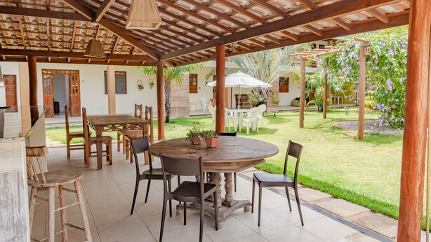 Uma área de varanda coberta com mesa e cadeiras de madeira redondas, cercada por um jardim com palmeiras e vista para uma casa.