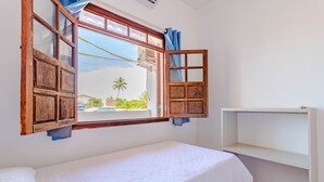 Um quarto com cama, janela com persianas de madeira e uma prateleira branca.