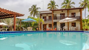 Piscina com espreguiçadeiras e guarda-sóis, rodeada por um edifício de dois andares com varandas, palmeiras e um espaço coberto de varanda.