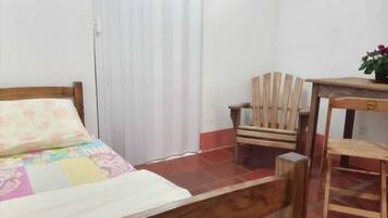 Um quarto pequeno com uma cama, uma cadeira de madeira, uma mesinha e um vaso com planta.