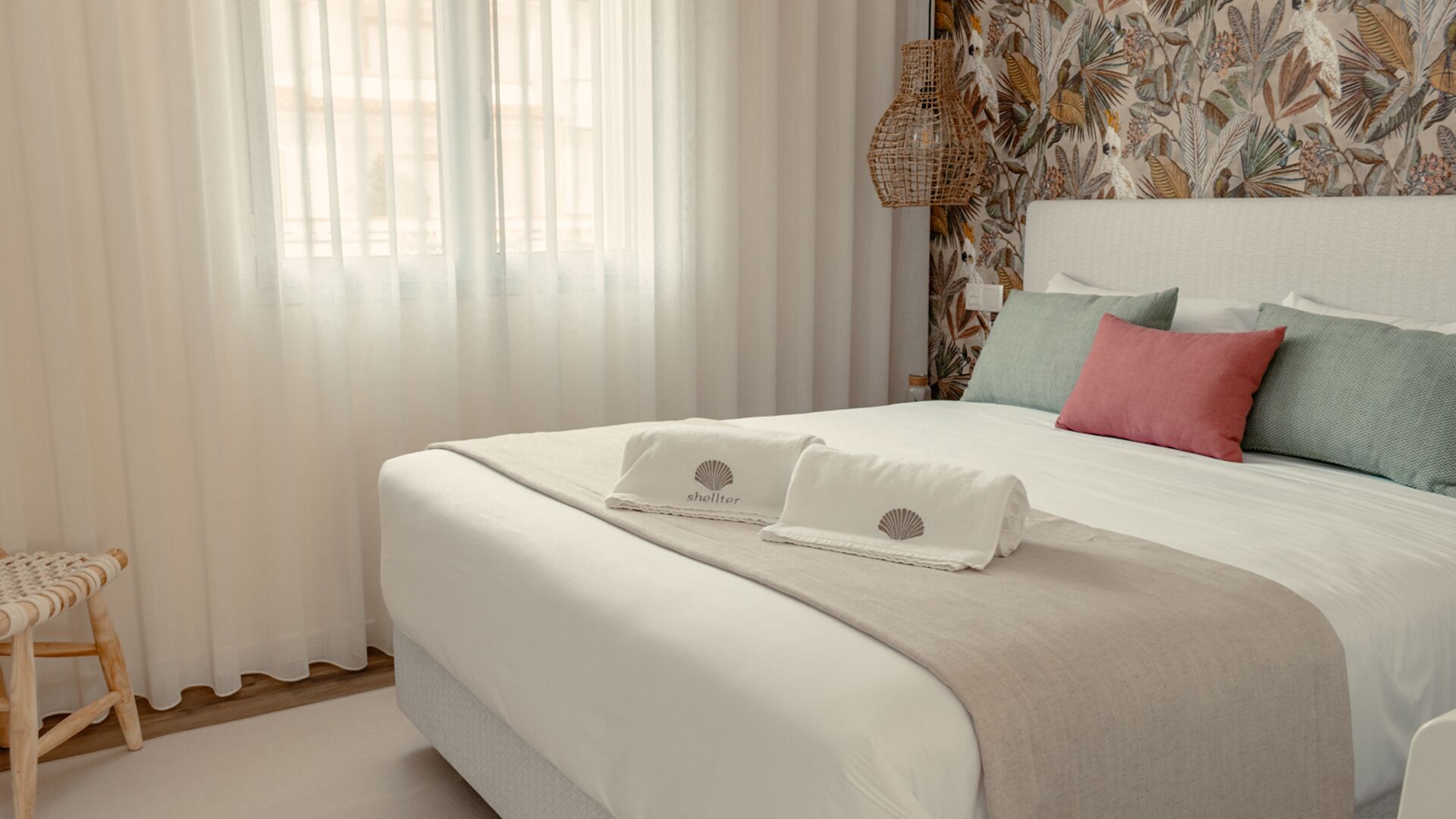 Uma cama bem arrumada com um cobreleito branco, duas almofadas com o logotipo de uma concha e um pequeno banquinho de madeira.