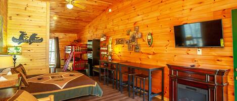 Interior acolhedor de uma cabana com teto de madeira, lareira, televisão e área de jantar.