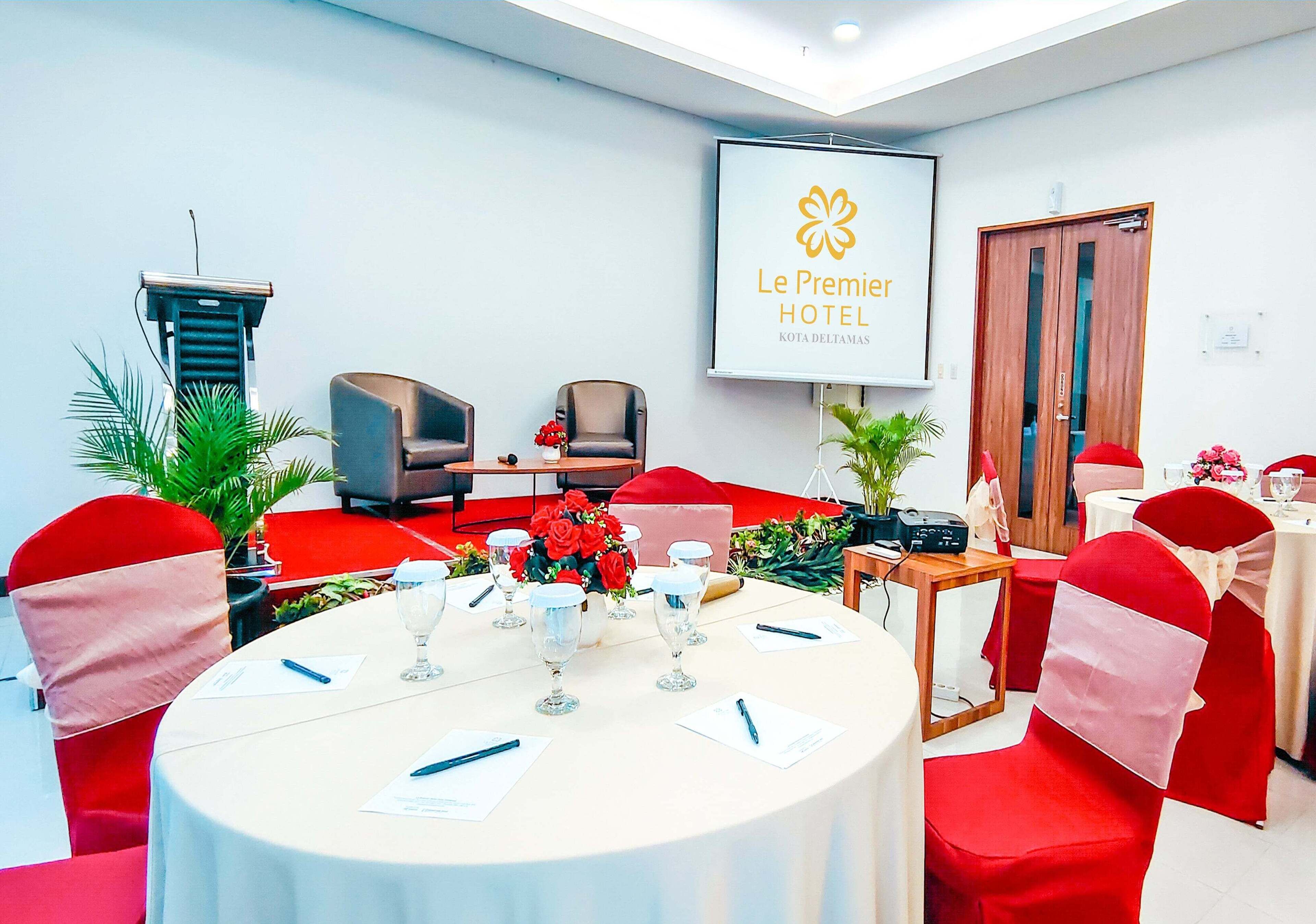 Uma sala de conferências com uma mesa branca, cadeiras vermelhas, uma tela de projetor e um tapete vermelho.