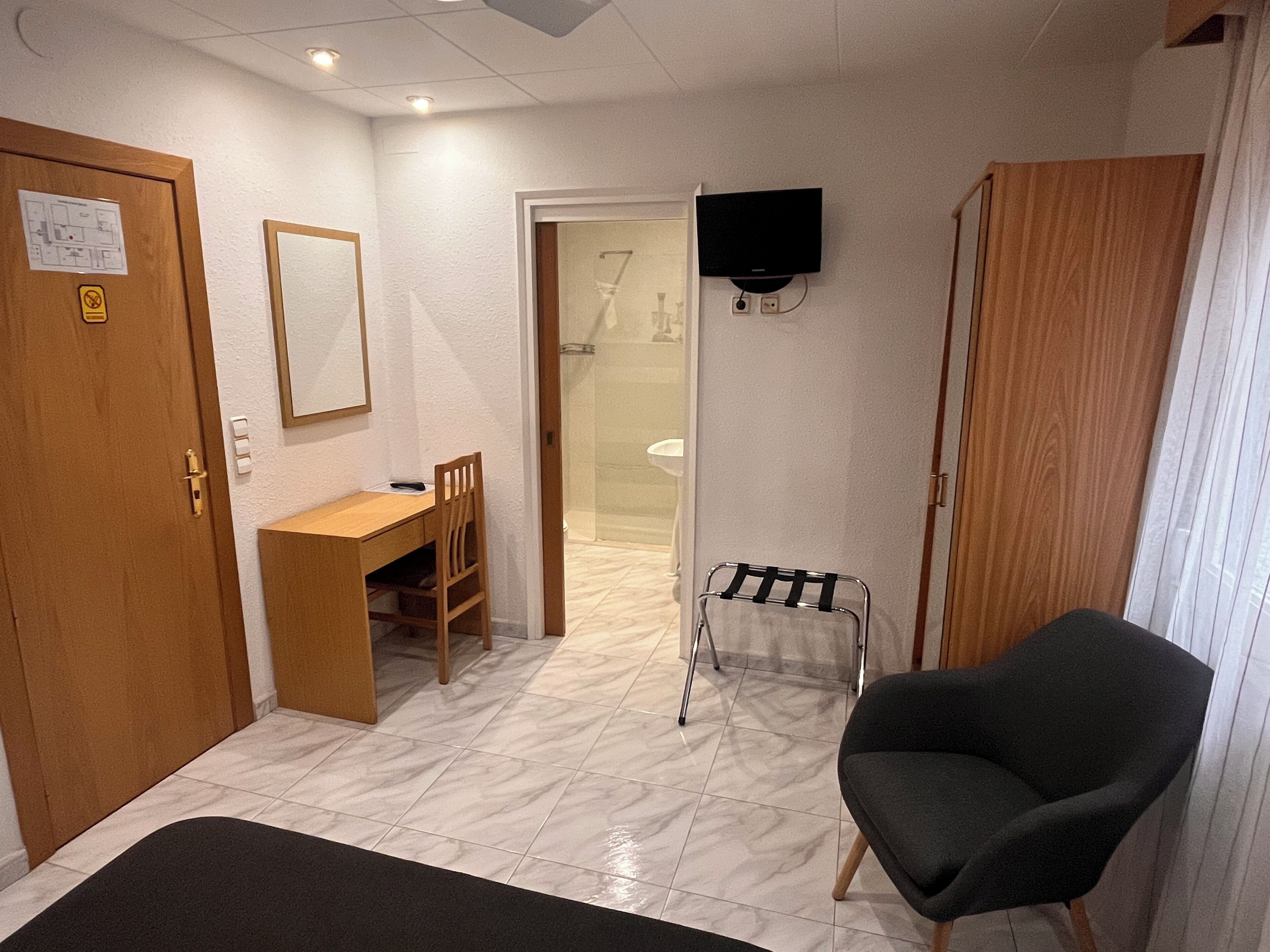 Quarto com escrivaninha de madeira, cadeira preta, televisão fixada na parede e área de chuveiro visível por uma porta aberta.