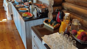 Um buffet de café da manhã com diversos alimentos e bebidas, ambientado em uma cabana de tronco com paredes e piso de madeira.