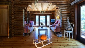 Interior de uma cabana de madeira com soalho de madeira, dois sofás com almofadas estampadas, uma mesa pequena com uma abóbora e uma cadeira branca.
