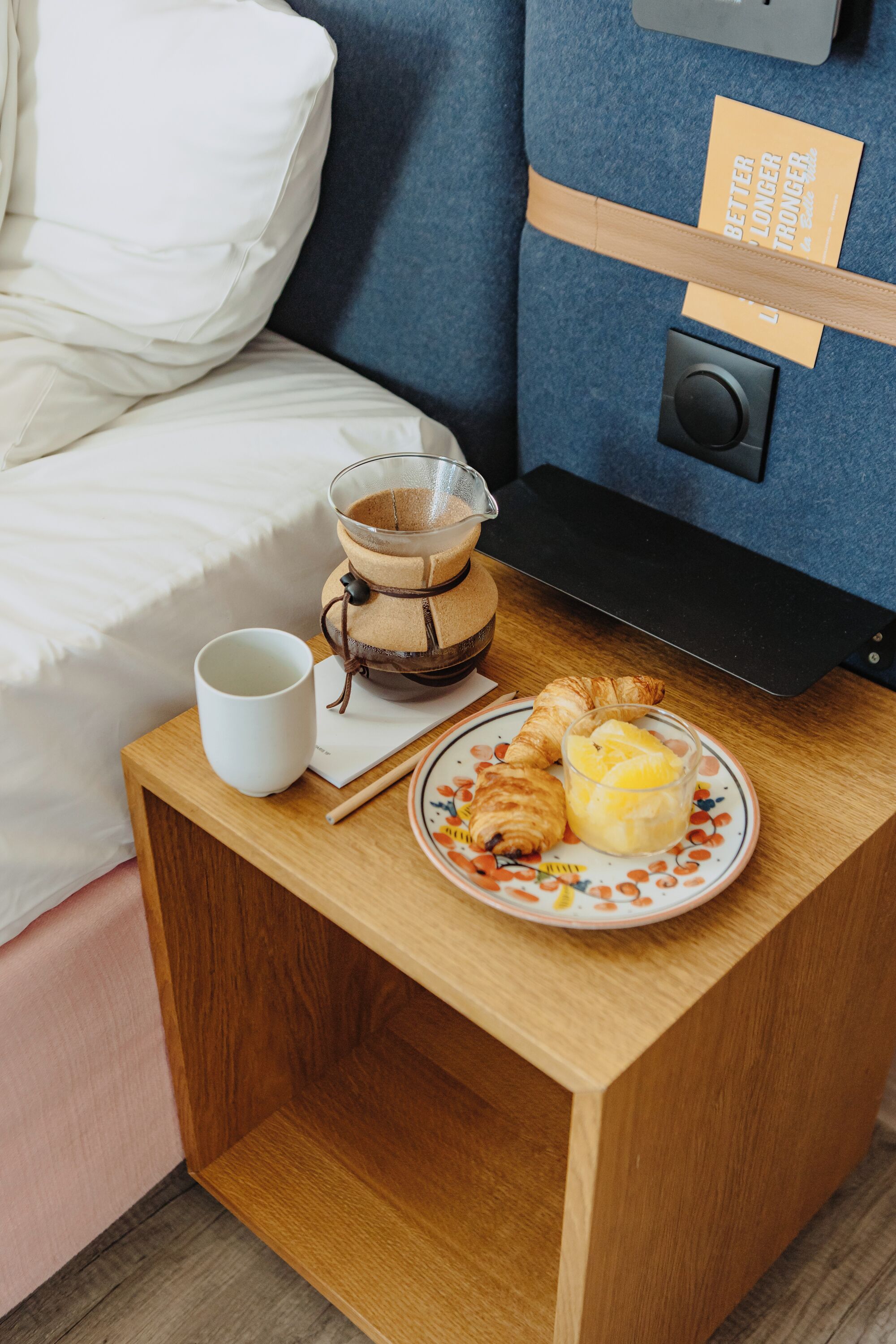 Cama bem arrumada com lençóis brancos, mesa de cabeceira com cafeteira, xícara e prato com croissants e frutas.