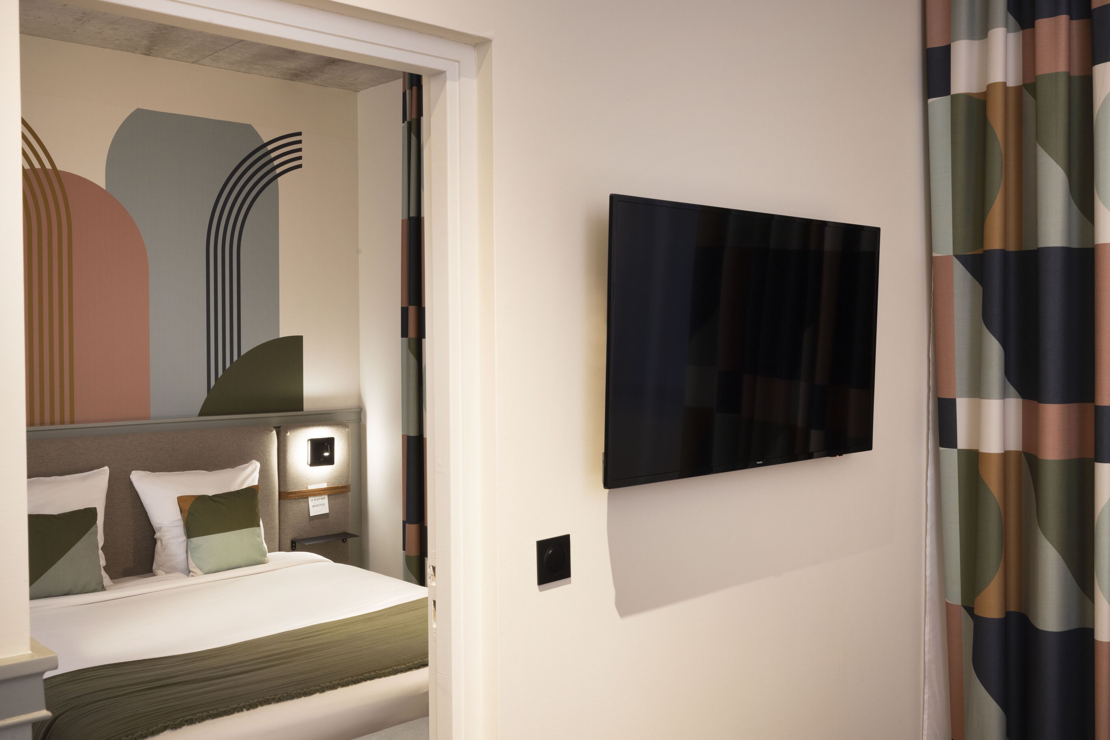 Quarto de hotel com cama, televisão fixada na parede e uma pintura abstrata moderna.