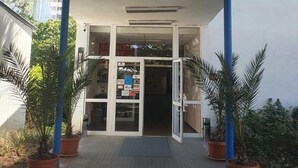 Entrada de um edifício com varanda coberta, plantas em vasos e vista clara do interior através de portas de vidro.