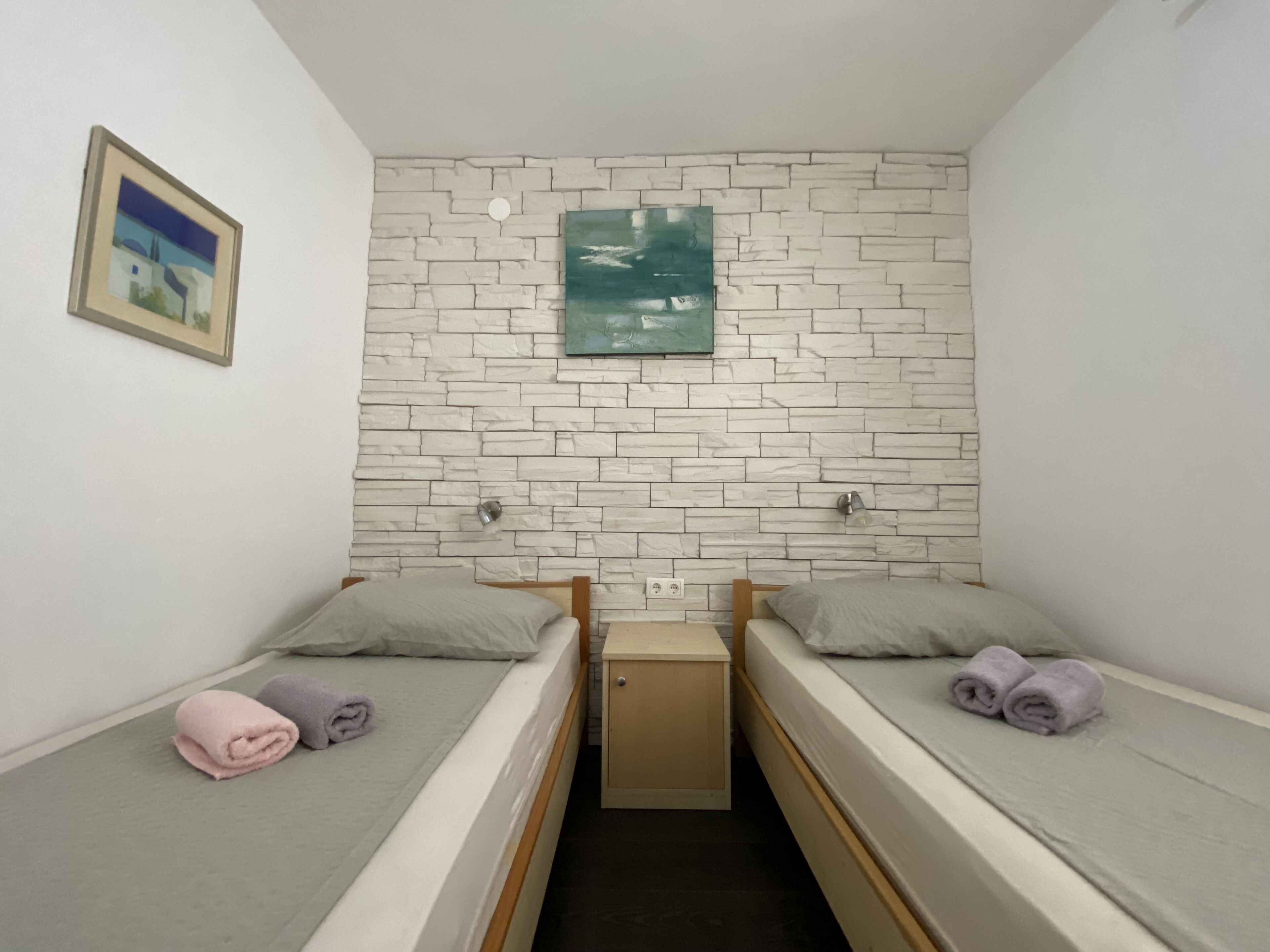 Quarto pequeno com duas camas de solteiro, uma mesa de cabeceira de madeira, uma luminária na parede e um quadro emoldurado.