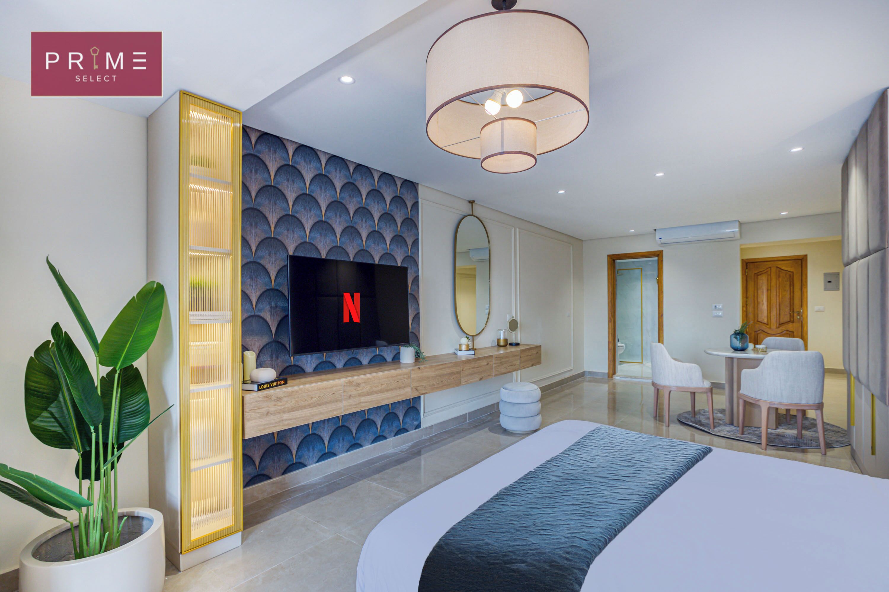 Quarto de hotel moderno com uma cama grande, uma televisão na parede, área de estar com duas cadeiras e uma planta em vaso.