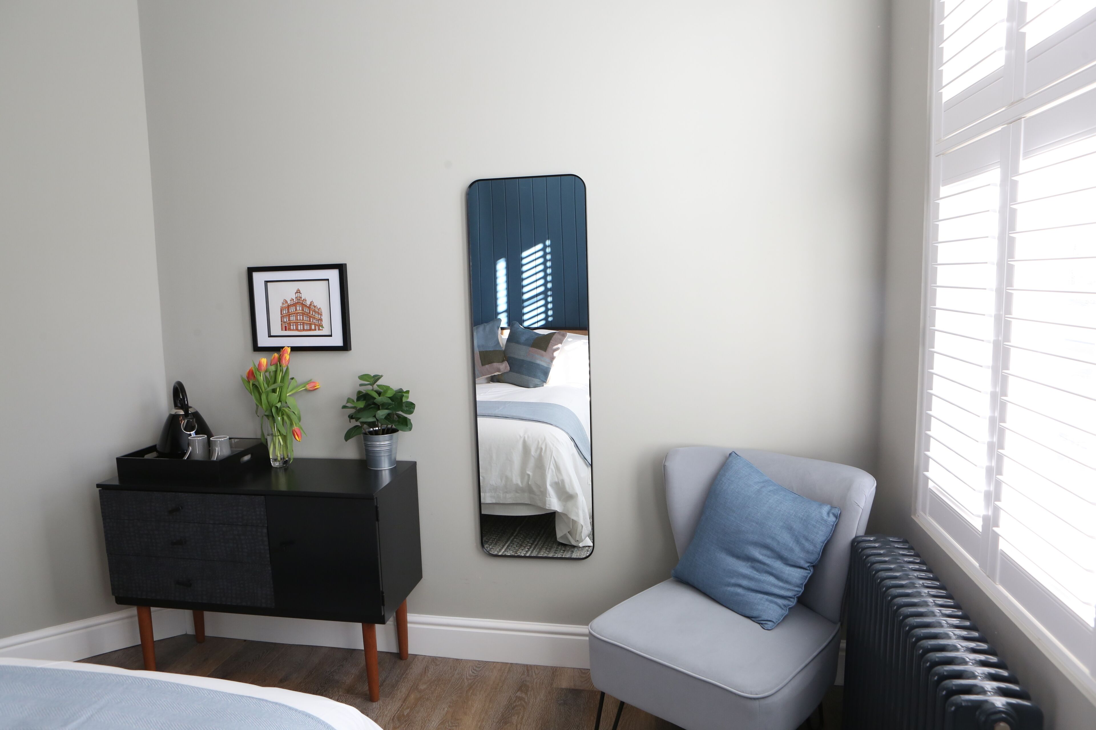 Quarto com cama, cómoda, espelho, radiador e janela com persianas.