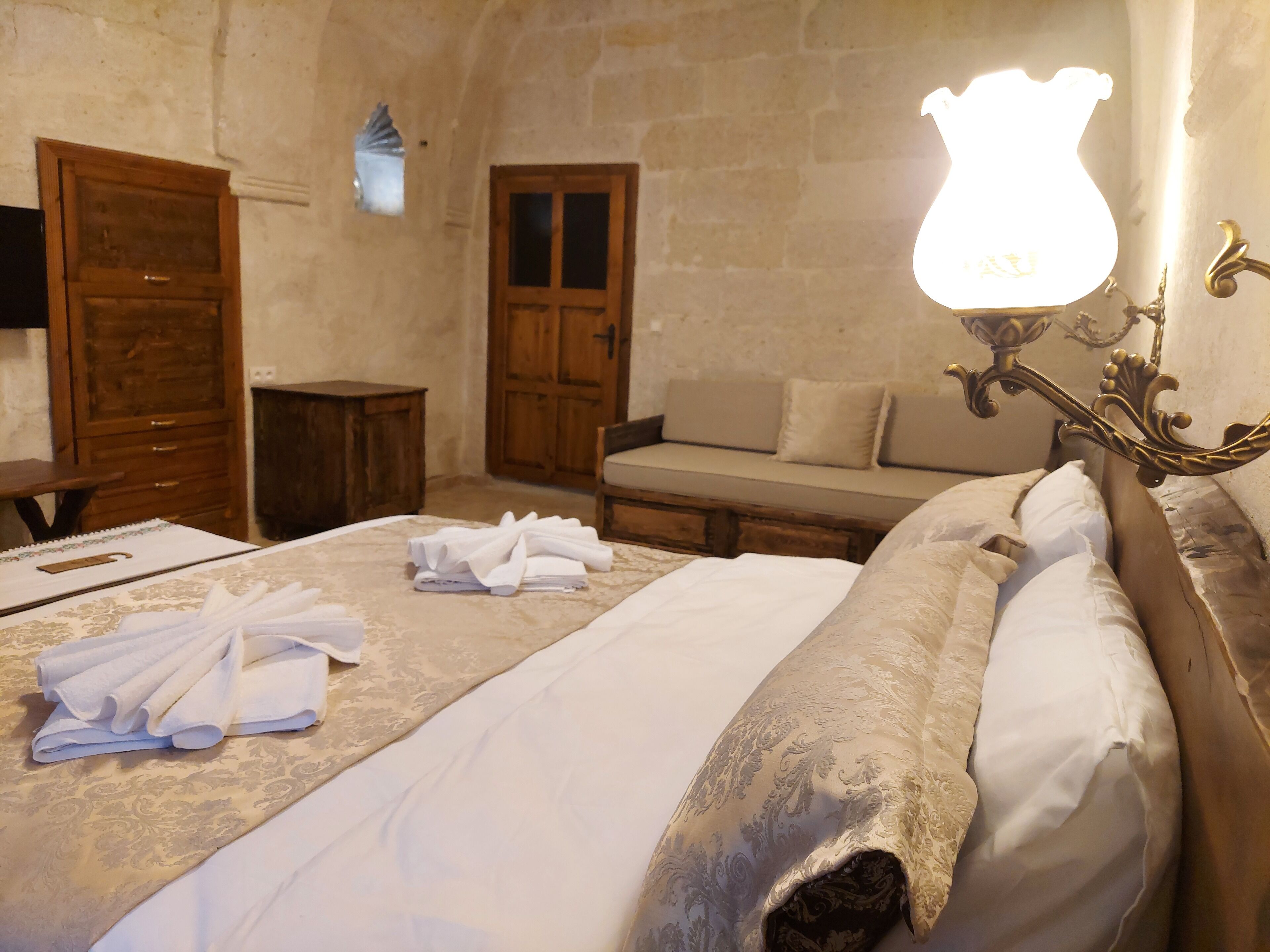 Quarto de hotel com uma cama, um banco de madeira, uma mesinha e uma luminária fixada na parede.