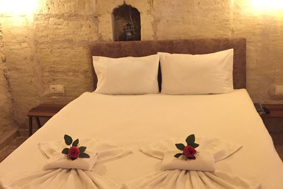 Quarto de hotel com uma cama grande, duas almofadas brancas e cabeceira de madeira. Há duas mesas de cabeceira com abajures, e uma parede de pedra com uma pequena janela.