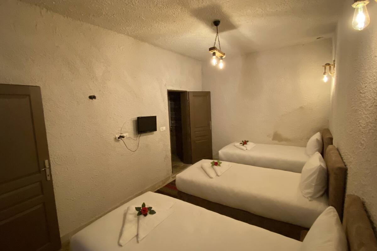 Quarto com duas camas, porta identificada com o número 6, televisão fixada na parede e luminária no teto.