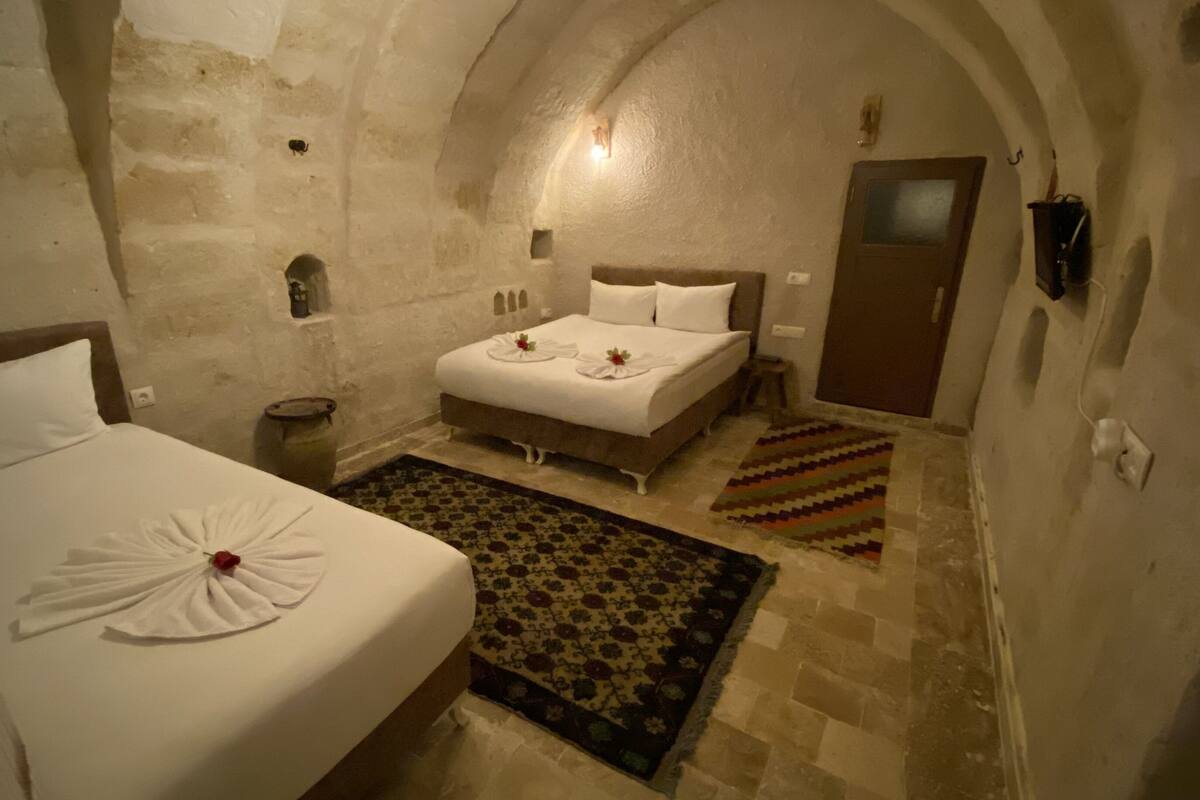 Quarto com uma cama, uma cama menor, uma mesinha de cabeceira, um tapete e uma porta.