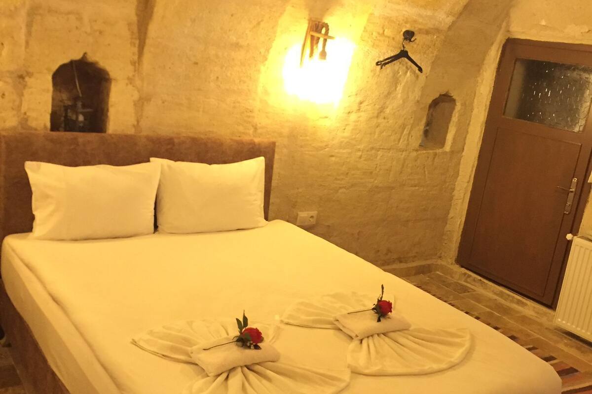 Quarto de hotel com uma cama grande, cabeceira de madeira, parede de pedra, uma janela pequena e uma porta.