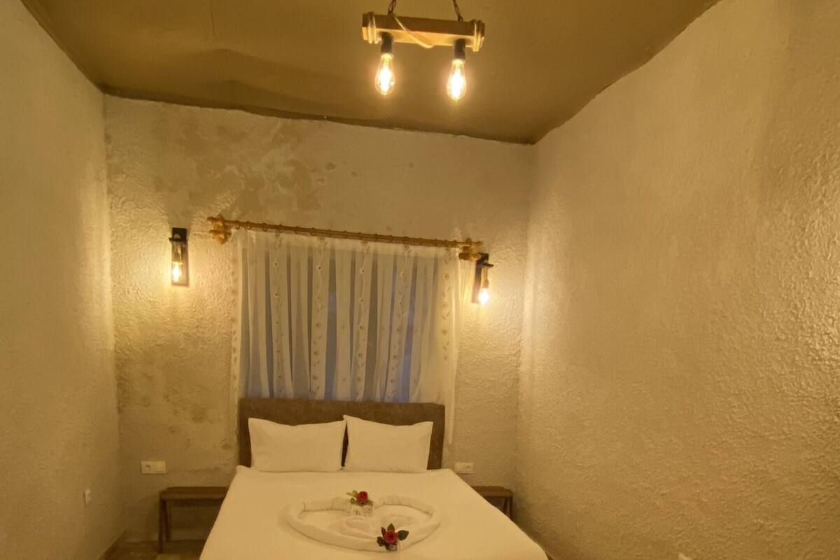 Quarto com cama, tapete estampado, luminária pendente e janela com cortinas.