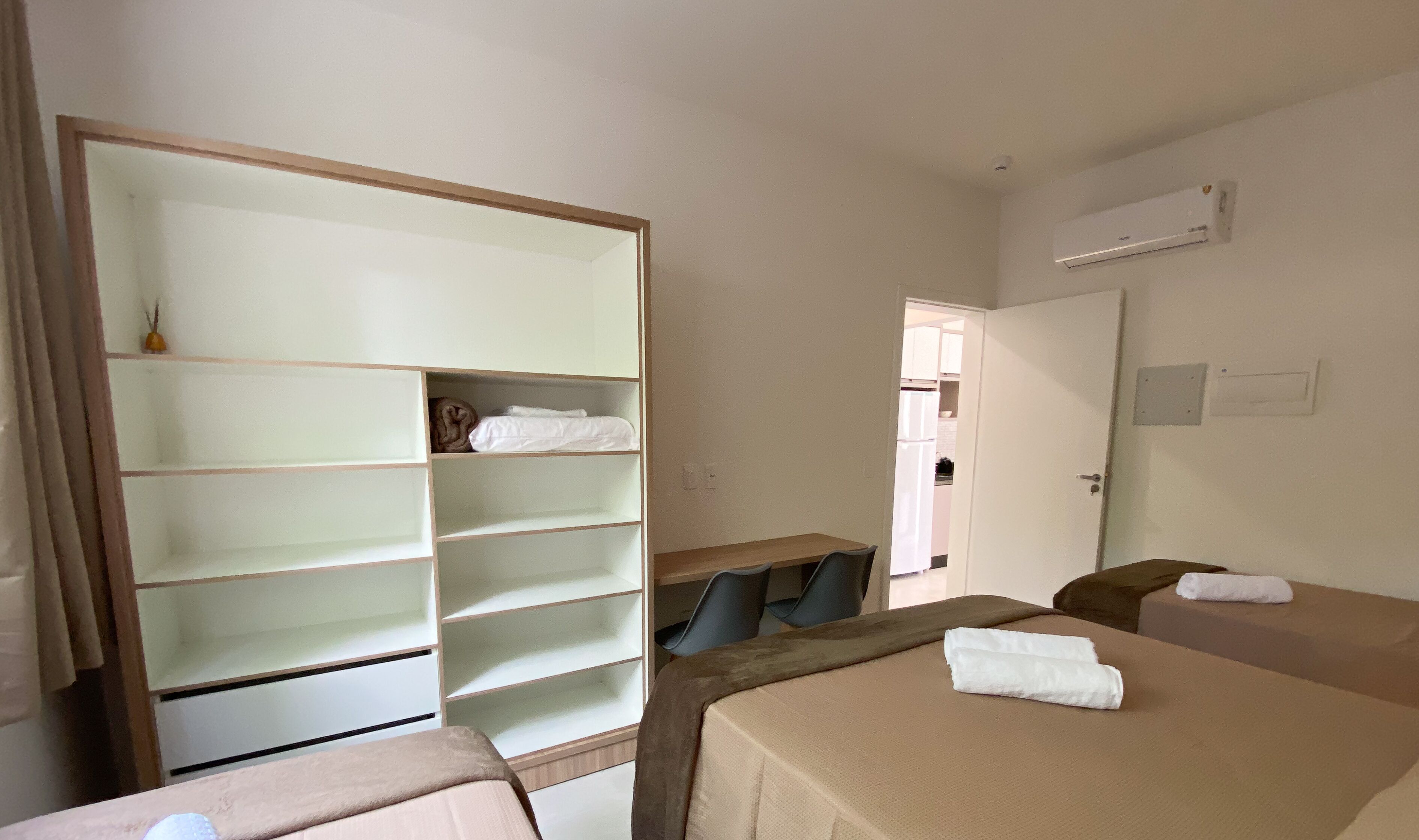 Quarto de hotel com um guarda-roupa espaçoso, uma cama com cobertura bege e uma escrivaninha com duas cadeiras.