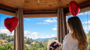 Mulher em roupão branco sentada em banheira com pétalas de rosas, segurando um buquê de flores, com vista para uma paisagem montanhosa através de janelas amplas e um candelabro acima.