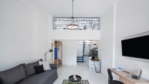 Uma sala de estar moderna com um sofá cinzento, uma mesa de centro com uma planta, uma área de jantar com uma mesa e cadeiras de madeira, e uma cozinha ao fundo.