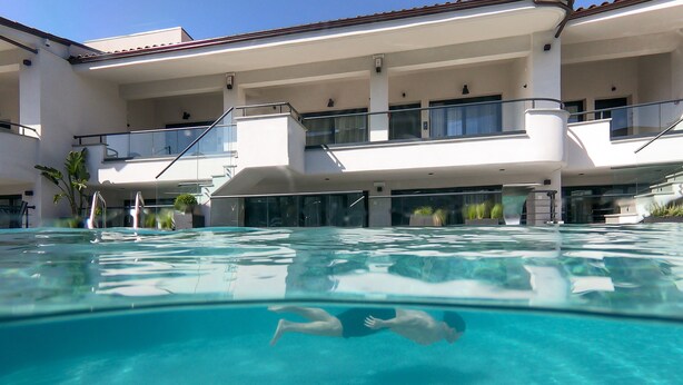 Uma piscina com água cristalina, rodeada por um edifício moderno com várias varandas e telhado em azulejo.