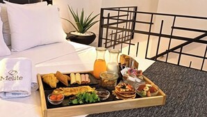 Cama bem arrumada com lençóis brancos e uma toalha dobrada com a etiqueta "Melite" ao lado. Uma bandeja com itens de café da manhã, incluindo pão, frutas e bebidas, está colocada sobre a cama.