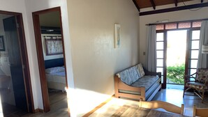 Quarto com um banco de madeira, sofá com almofadas estampadas, cama com colcha florida e vista para o verde através de grandes janelas.