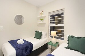 Quarto de hotel com duas camas, um espelho redondo, uma janela com persianas, uma mesa de cabeceira com abajur e uma pequena prateleira com uma planta.