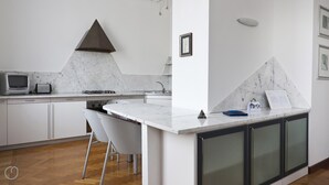 Cozinha moderna com bancada de mármore, fogão embutido, micro-ondas e pia. Possui armários brancos e um espaço de jantar com mesa e cadeiras.