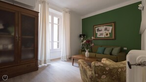 Uma sala de estar com parede verde, um armário de madeira, um sofá floral, uma mesa de centro com um vaso de flores e uma colagem de fotos emolduradas.