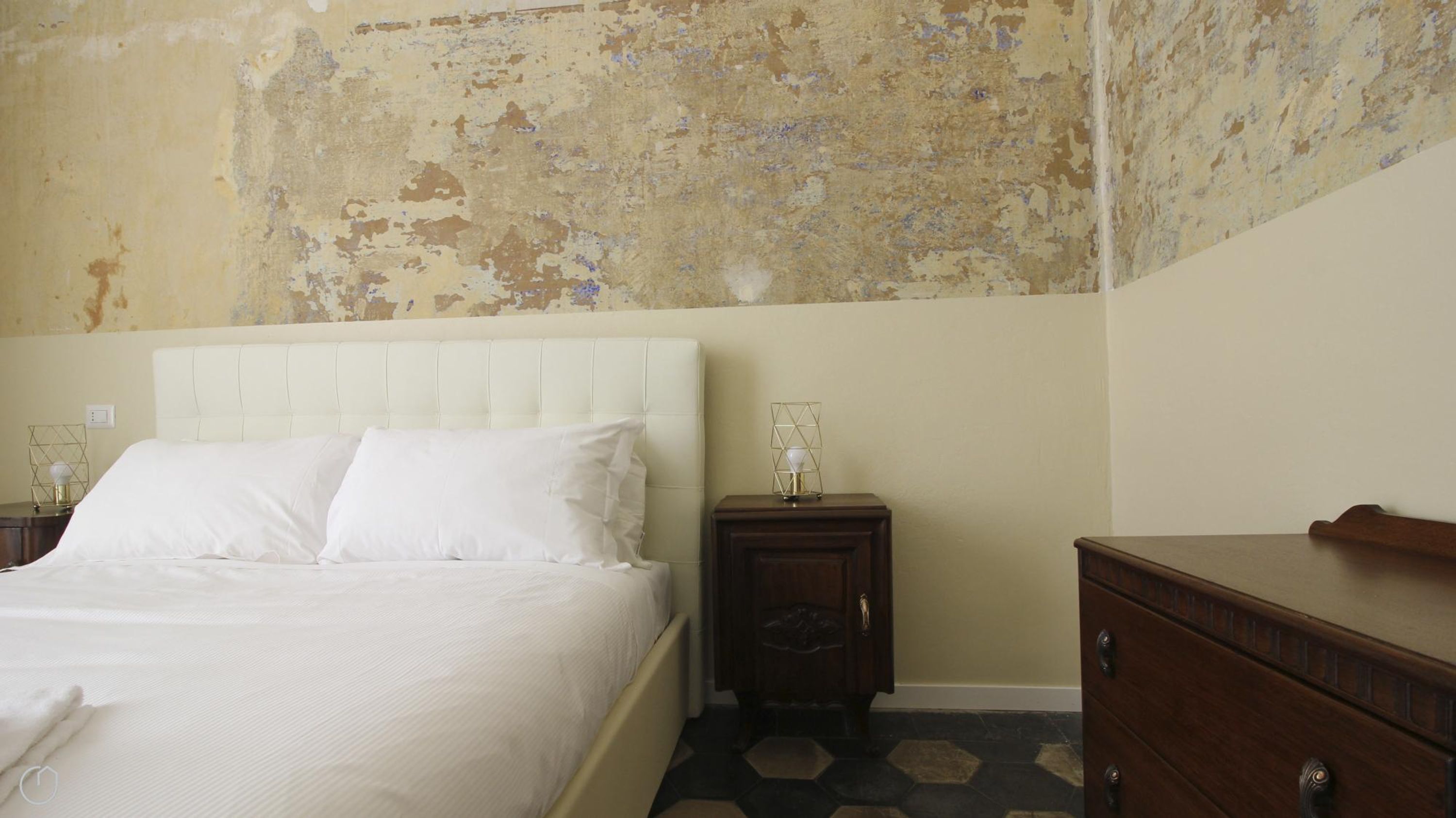 Quarto com uma cama grande, cômoda de madeira e mesa de cabeceira com abajur. As paredes têm textura e apresentam sinais de uso.