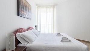 Quarto de hotel com uma cama grande, roupa de cama branca, almofadas vermelhas estampadas, uma luminária branca e um quadro na parede.