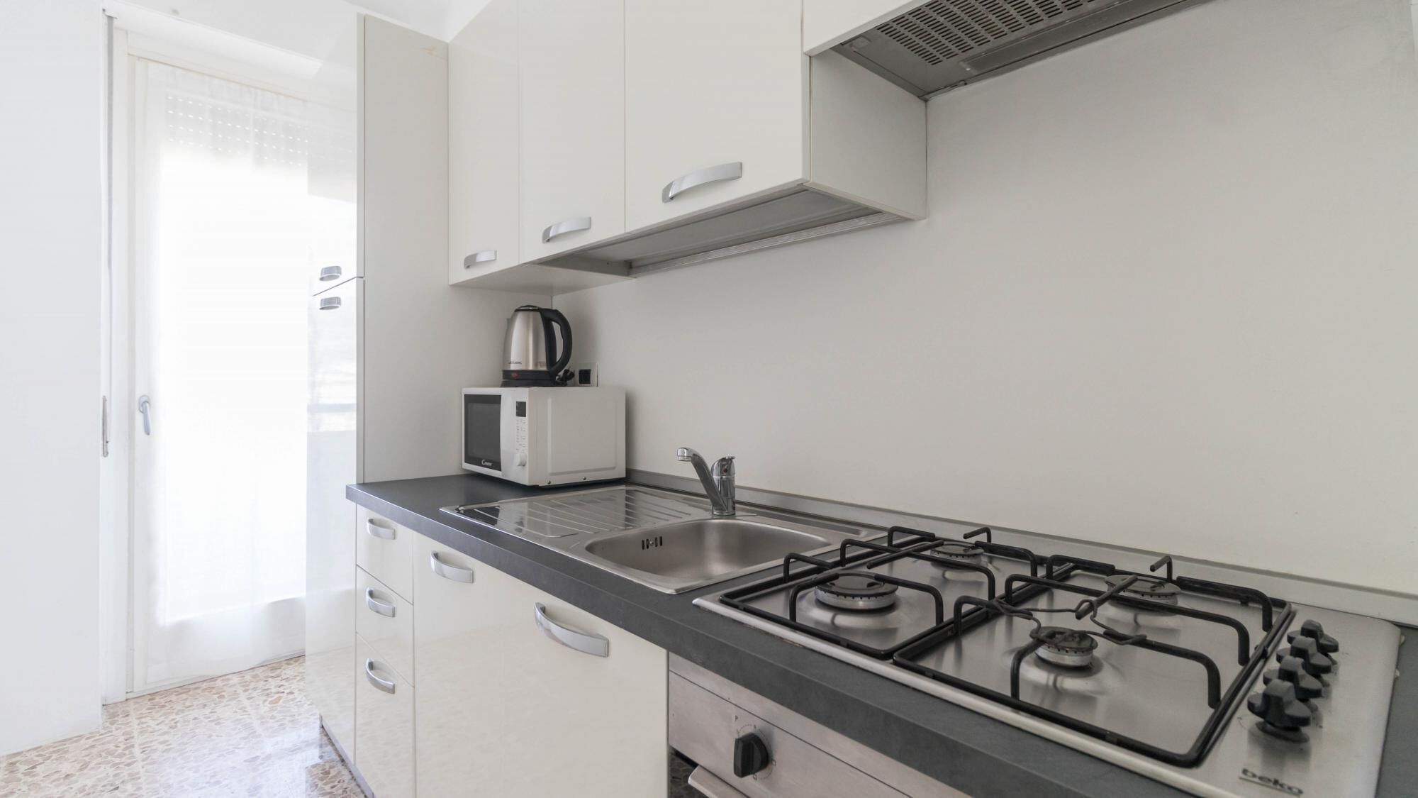 Uma cozinha moderna com armários brancos, uma pia de aço inoxidável, fogão a gás e micro-ondas.