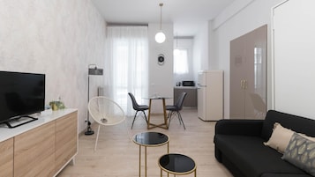Uma sala moderna com um sofá preto, uma televisão, uma área de refeições com cadeiras, uma mesa pequena e uma cozinha ao fundo.