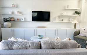 Uma sala de estar moderna com uma televisão de tela plana, prateleiras brancas com objetos decorativos, uma mesa redonda com uma tigela e um sofá cinza.