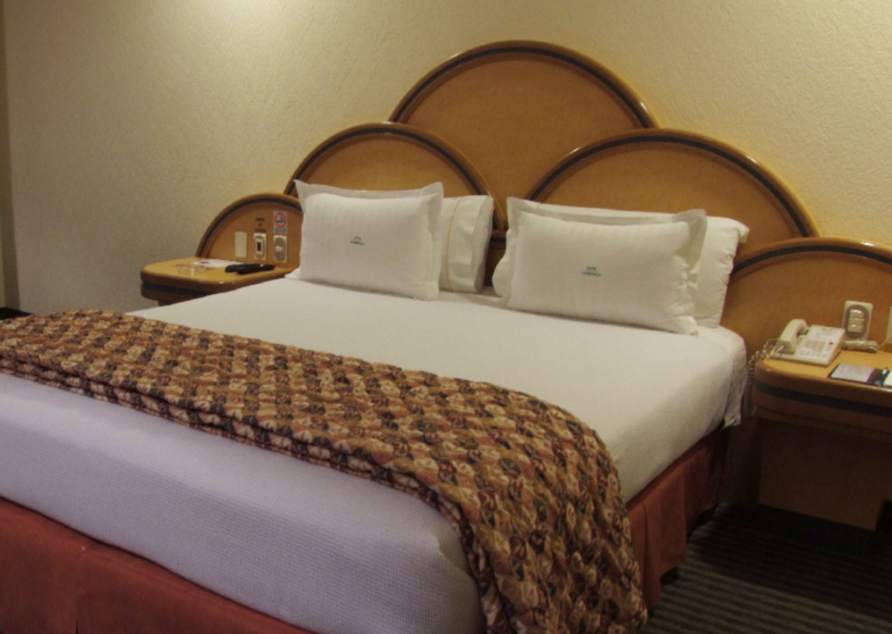 Quarto de hotel com uma cama grande, cabeceira de madeira, mesa de cabeceira com telefone e manta decorativa.