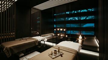 Uma sala de spa moderna com duas macas de massagem, um balcão de recepção e uma grande janela com vista para a cidade.