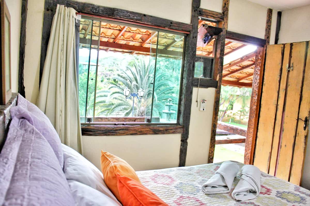 Quarto com cama, janela com cortinas, porta e vista para o verde lá fora.