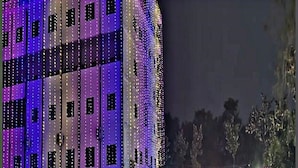 Um edifício com vários andares, cada um iluminado com luzes coloridas. Árvores e uma fonte estão em primeiro plano.