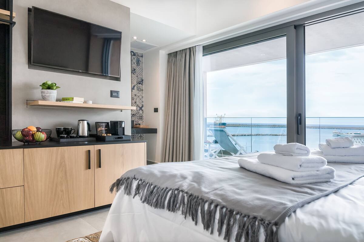 Quarto de hotel moderno com uma cama grande, televisão de tela plana na parede, pequena cozinha com cafeteira e varanda com vista para o mar.