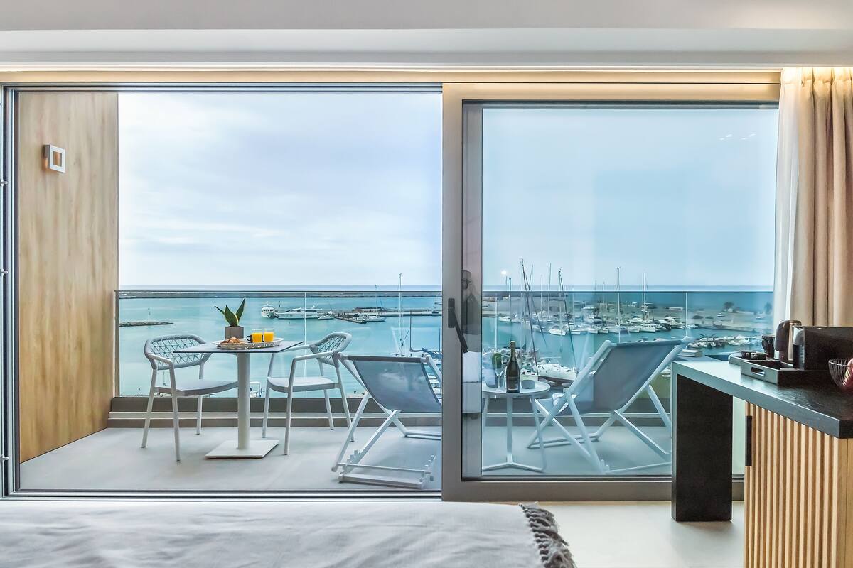 Uma varanda com vista para o porto de iates, uma mesa de jantar com cadeiras e uma pequena mesa com garrafa e copos.