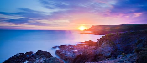 Paisagem costeira com terreno rochoso e um pôr do sol sobre o oceano.