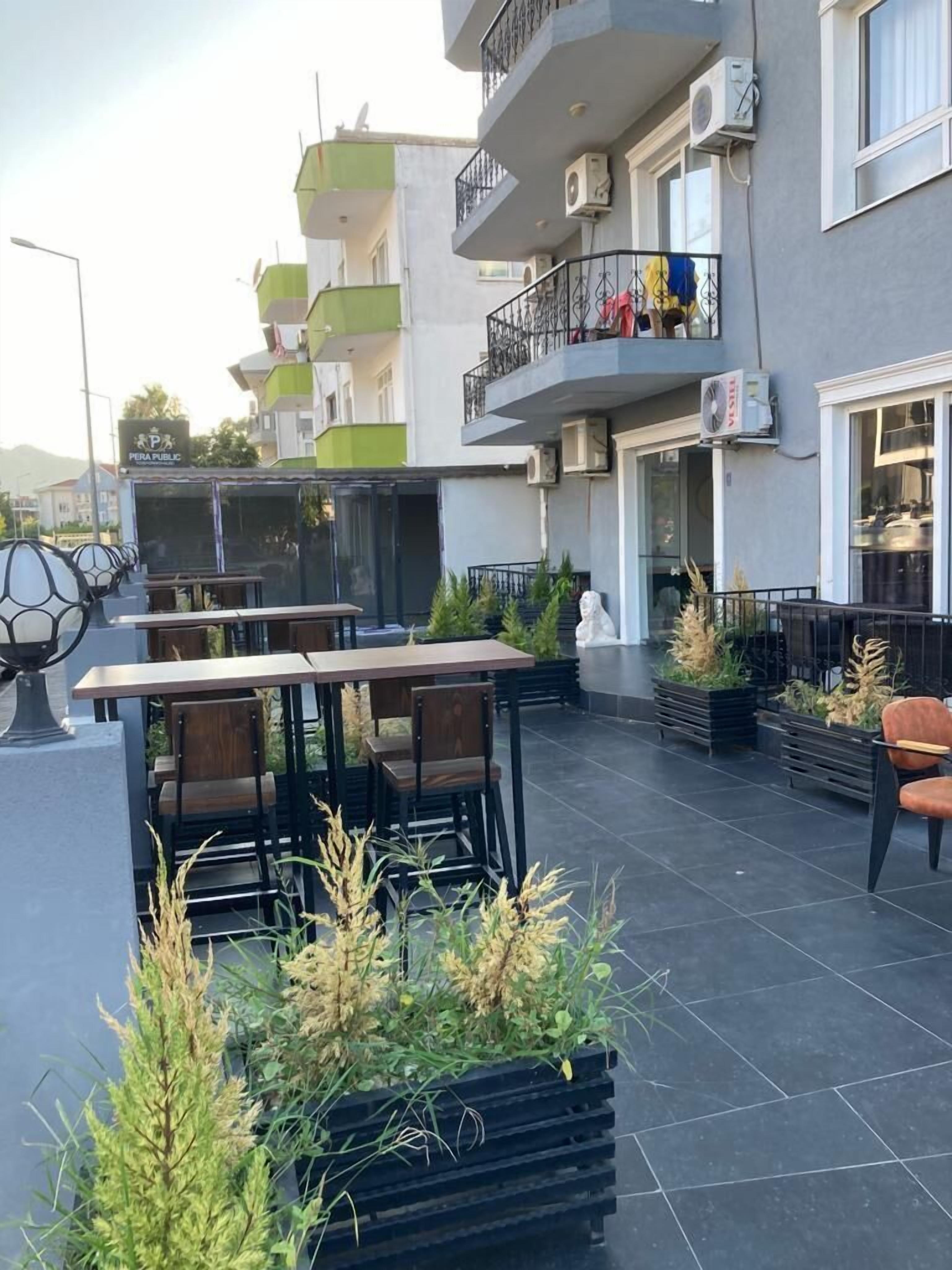 Uma área de pátio externa com mesas e cadeiras, rodeada por plantas em vasos e unidades de ar condicionado no edifício.