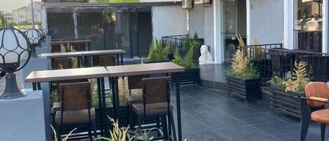 Uma área de pátio externa com mesas e cadeiras, rodeada por plantas em vasos e unidades de ar condicionado no edifício.