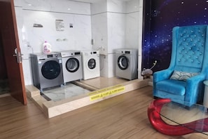 Quarto de hotel com máquina de lavar, uma poltrona azul e uma mesa de centro de vidro.