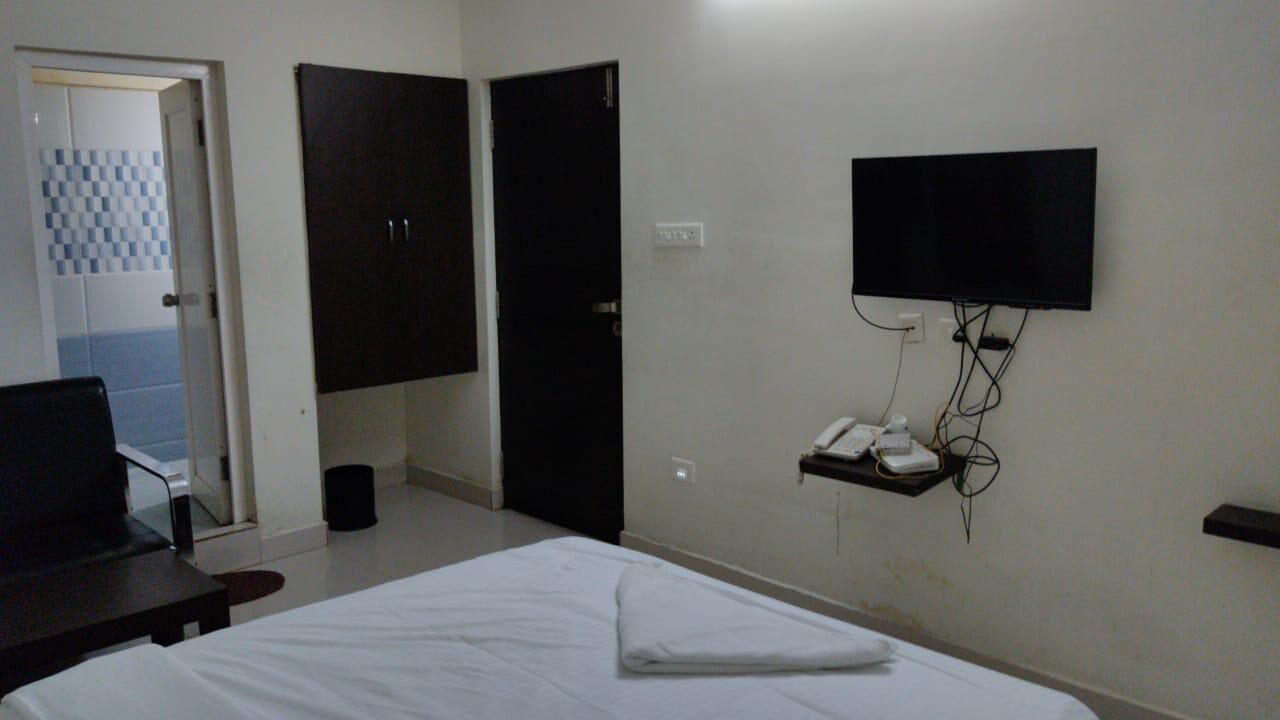 Quarto de hotel com cama, escrivaninha, cadeira, televisão fixada na parede e porta que dá para outro cômodo.