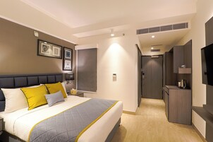Quarto de hotel moderno com uma cama grande, uma mesinha de cabeceira, televisão de tela plana e uma pequena cozinha.